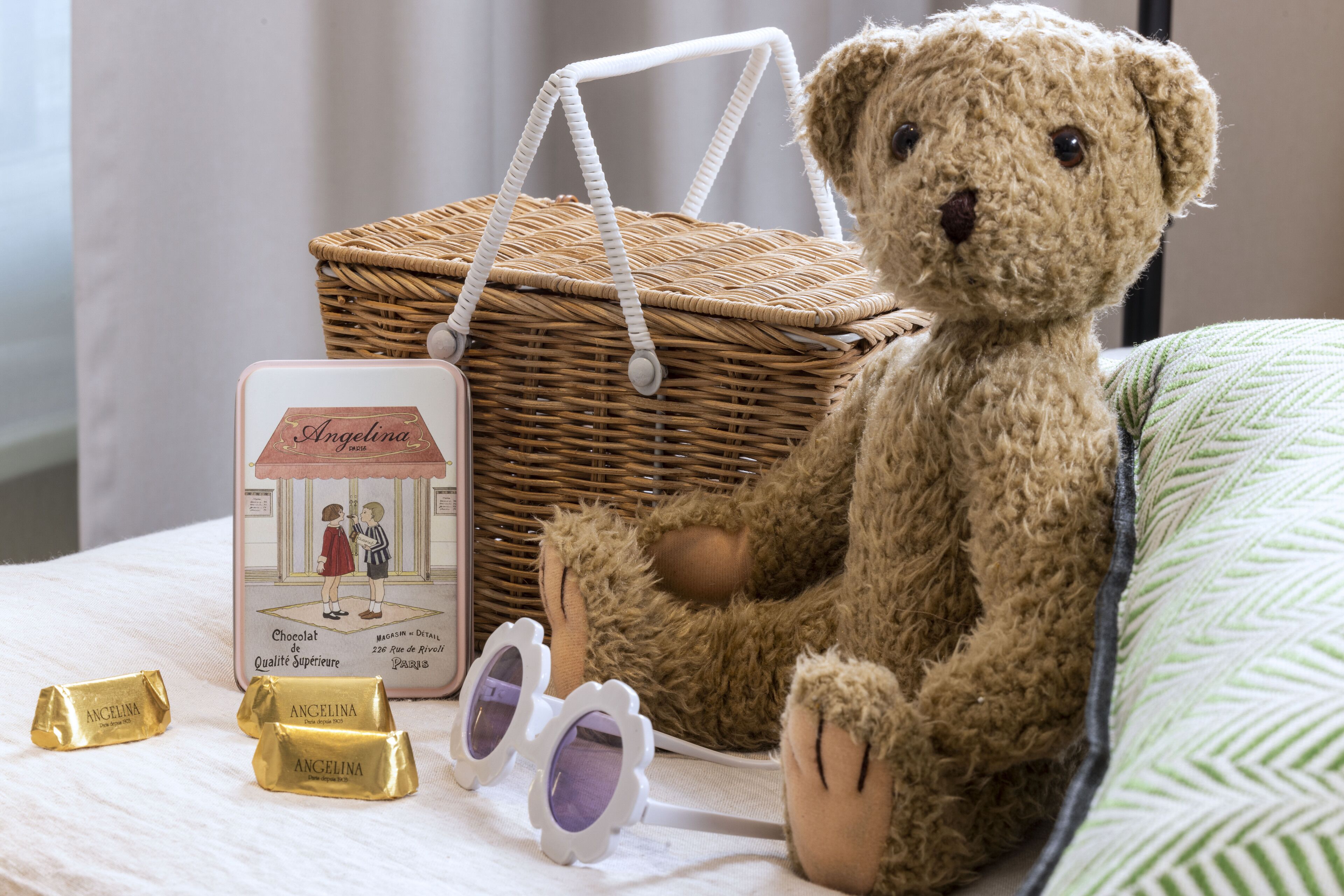 Um urso de pelúcia, uma cesta de vime, uma barra de chocolate e um par de óculos de sol sobre uma cama.