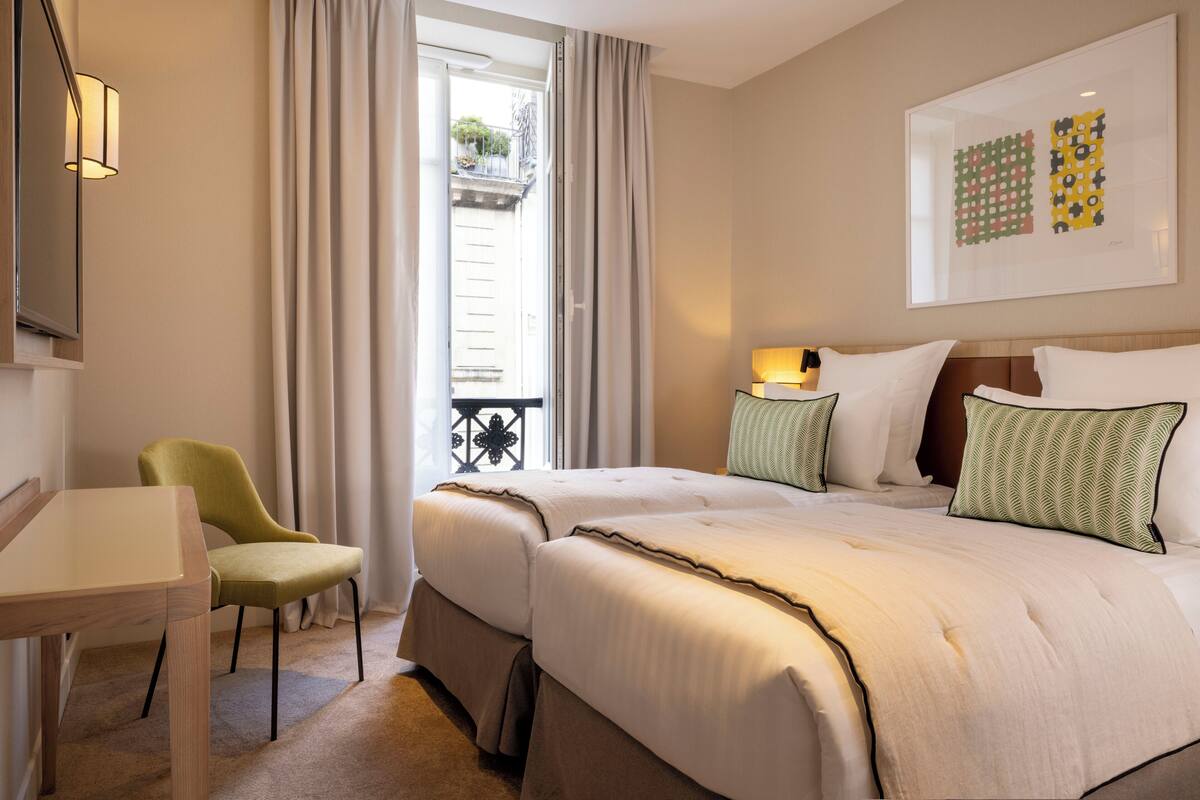 Quarto de hotel com duas camas, uma escrivaninha, uma cadeira e varanda com grade.