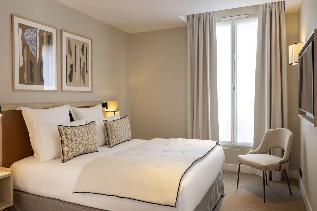 Quarto de hotel com uma cama grande, duas almofadas, uma cadeira, uma mesa de cabeceira, um abajur e duas molduras na parede.