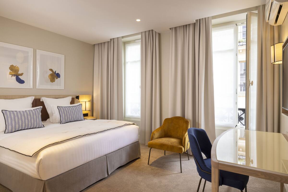Quarto de hotel com uma cama grande, uma escrivaninha com cadeira, uma mesa de cabeceira com luminária e uma janela com cortinas.