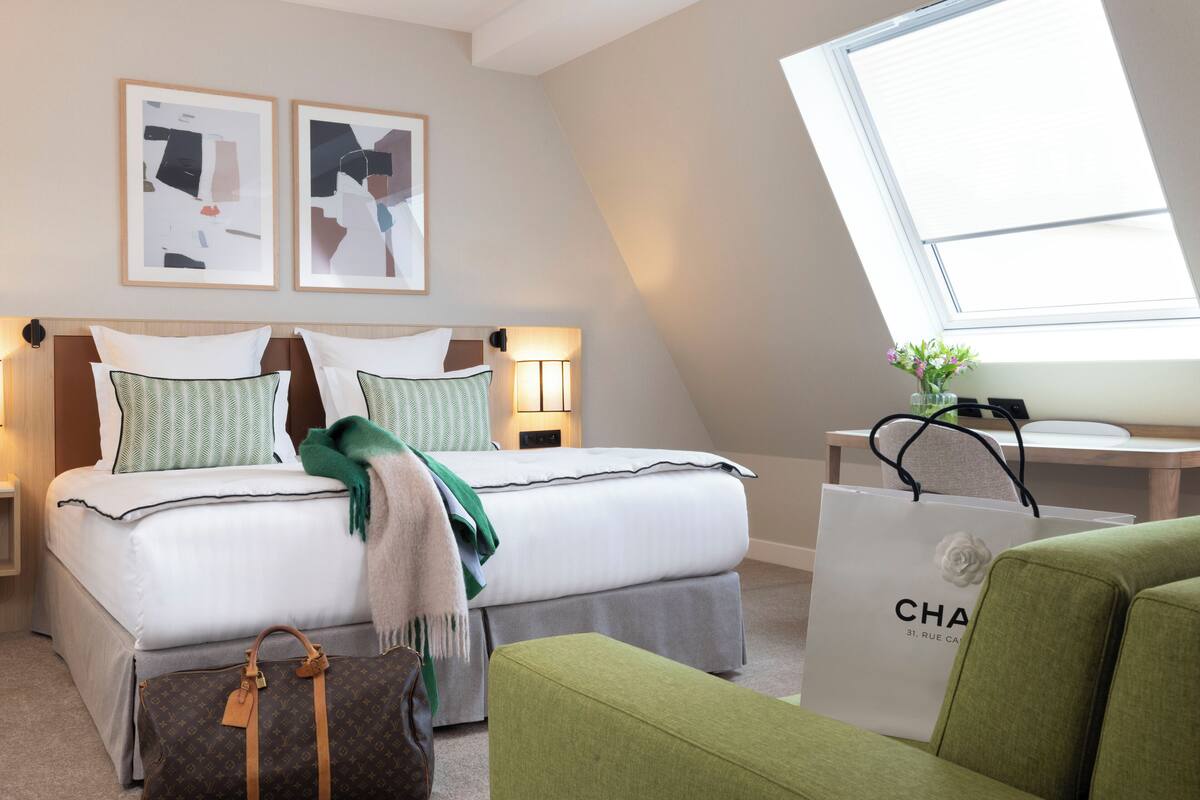 Quarto de hotel com cama, duas almofadas, abajur, cadeira verde, mala e janela com persianas.