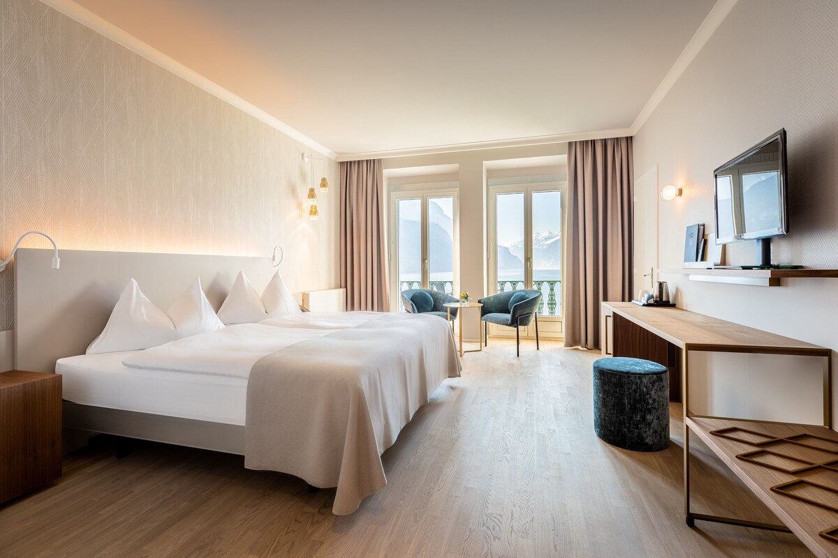 Quarto de hotel com cama, mesas de cabeceira, escrivaninha, cadeira, um banquinho redondo, televisão e varanda com cortinas.