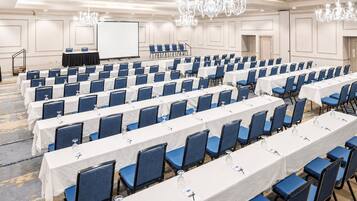 Uma sala de conferências com mesas e cadeiras dispostas em fileiras, uma tela de projetor e um palco.