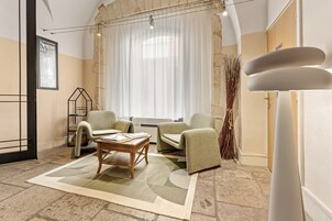 Quarto com duas poltronas, uma mesa de centro de madeira, tapete estampado, uma janela grande com cortinas translúcidas e um abajur moderno.