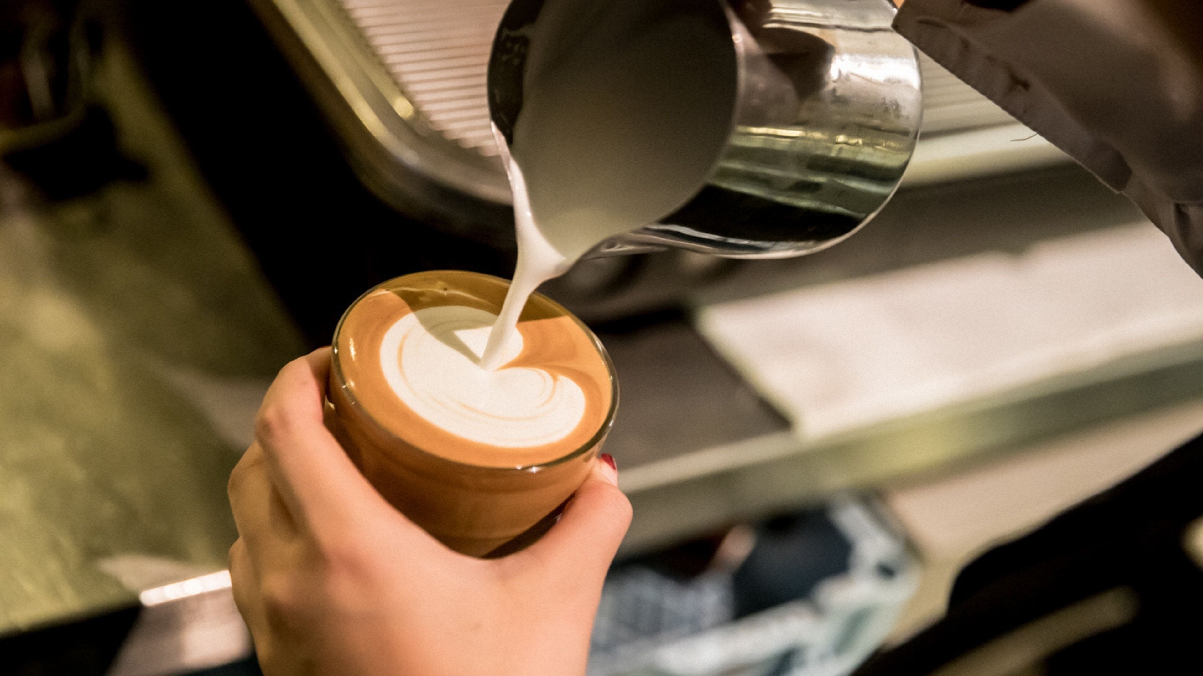 Alguém despejando leite numa chávena de café, formando um desenho em forma de coração na superfície.