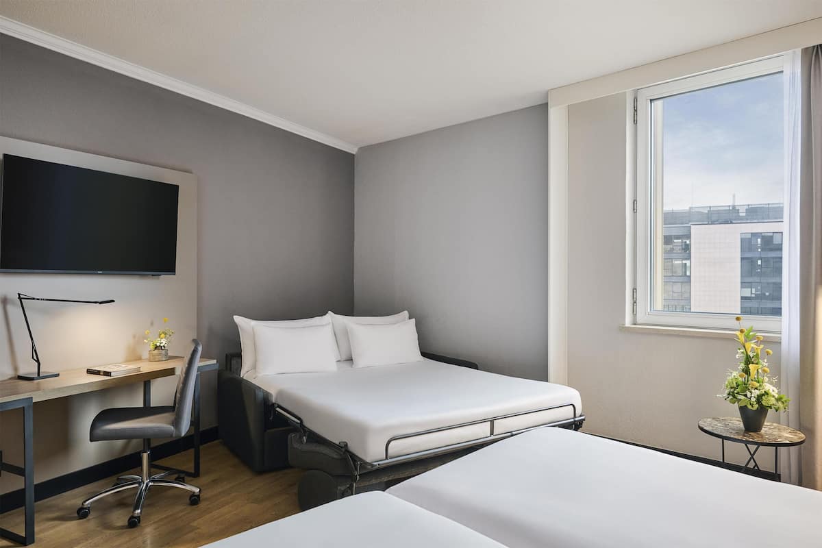 Quarto de hotel com cama, escrivaninha com cadeira, televisão na parede e janela com vista para edifícios.