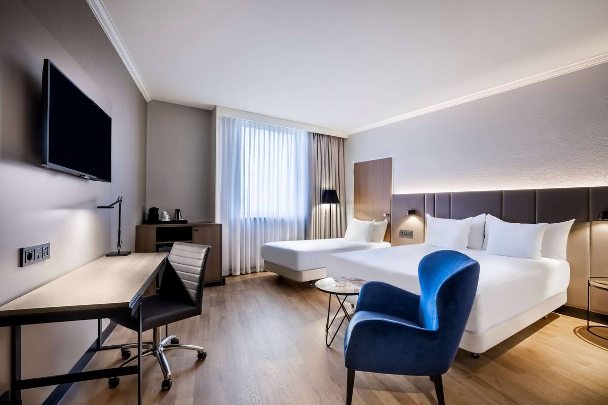 Quarto de hotel com cama, escrivaninha, cadeira e televisão.