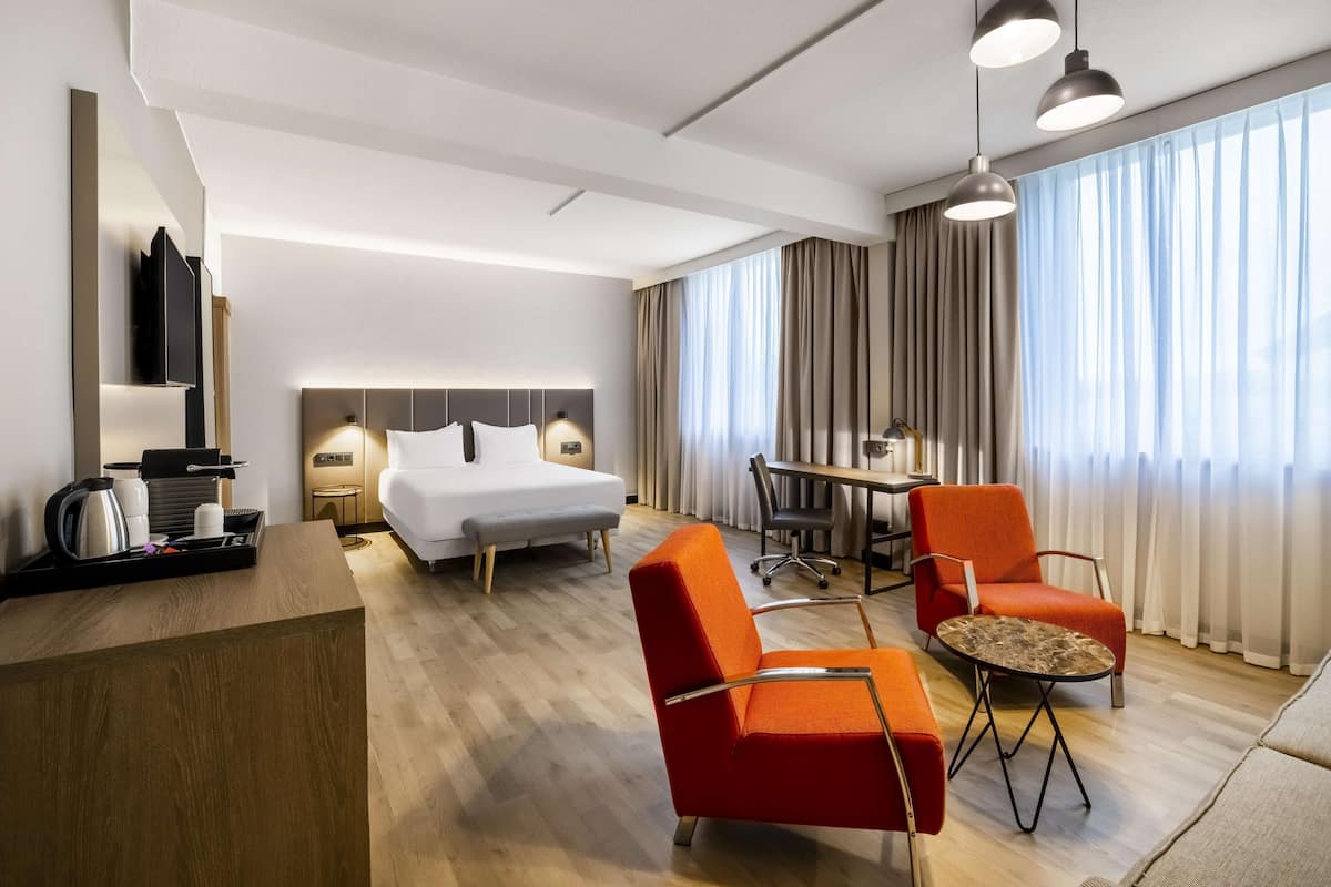 Quarto de hotel com cama, escrivaninha, duas cadeiras, mesa e uma janela grande com cortinas.