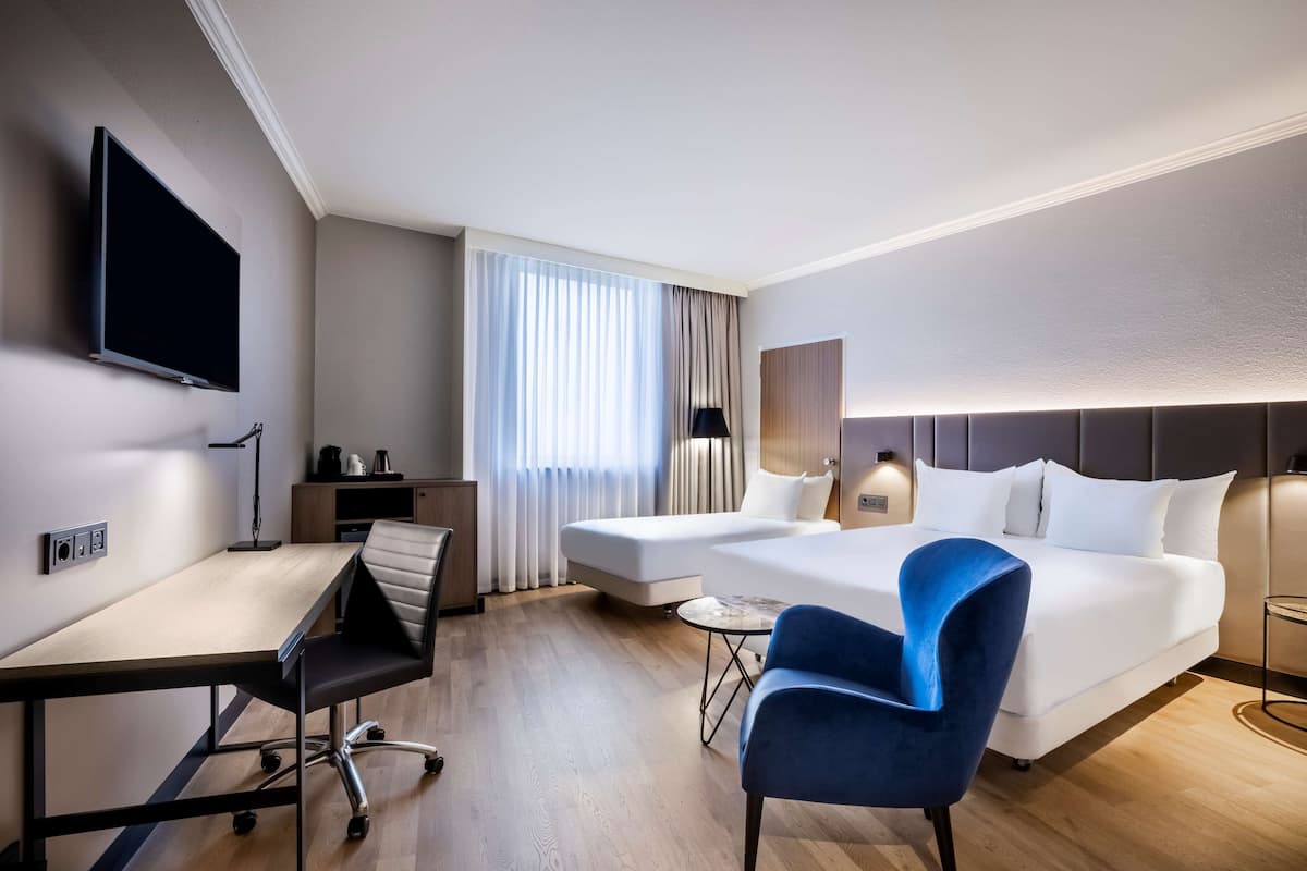 Quarto de hotel com cama, escrivaninha, cadeira, televisão e janela com cortinas.