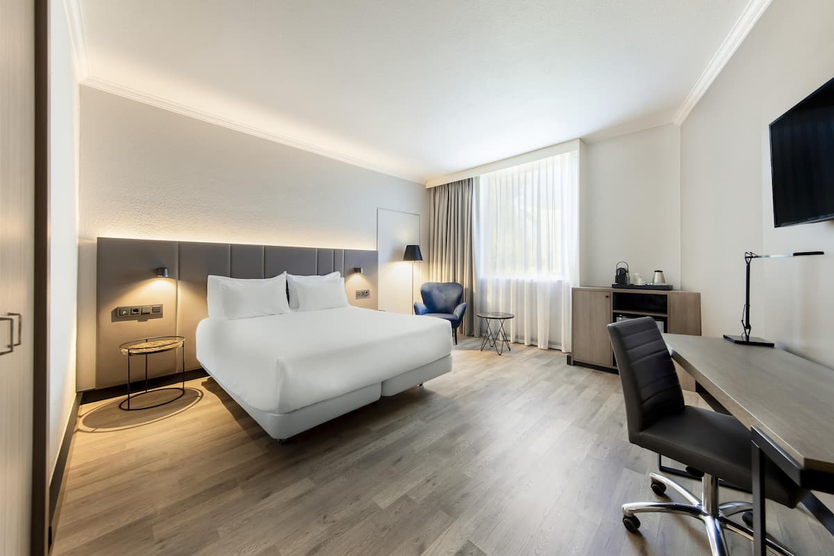 Quarto de hotel com cama, escrivaninha, cadeira, mesinha e televisão.