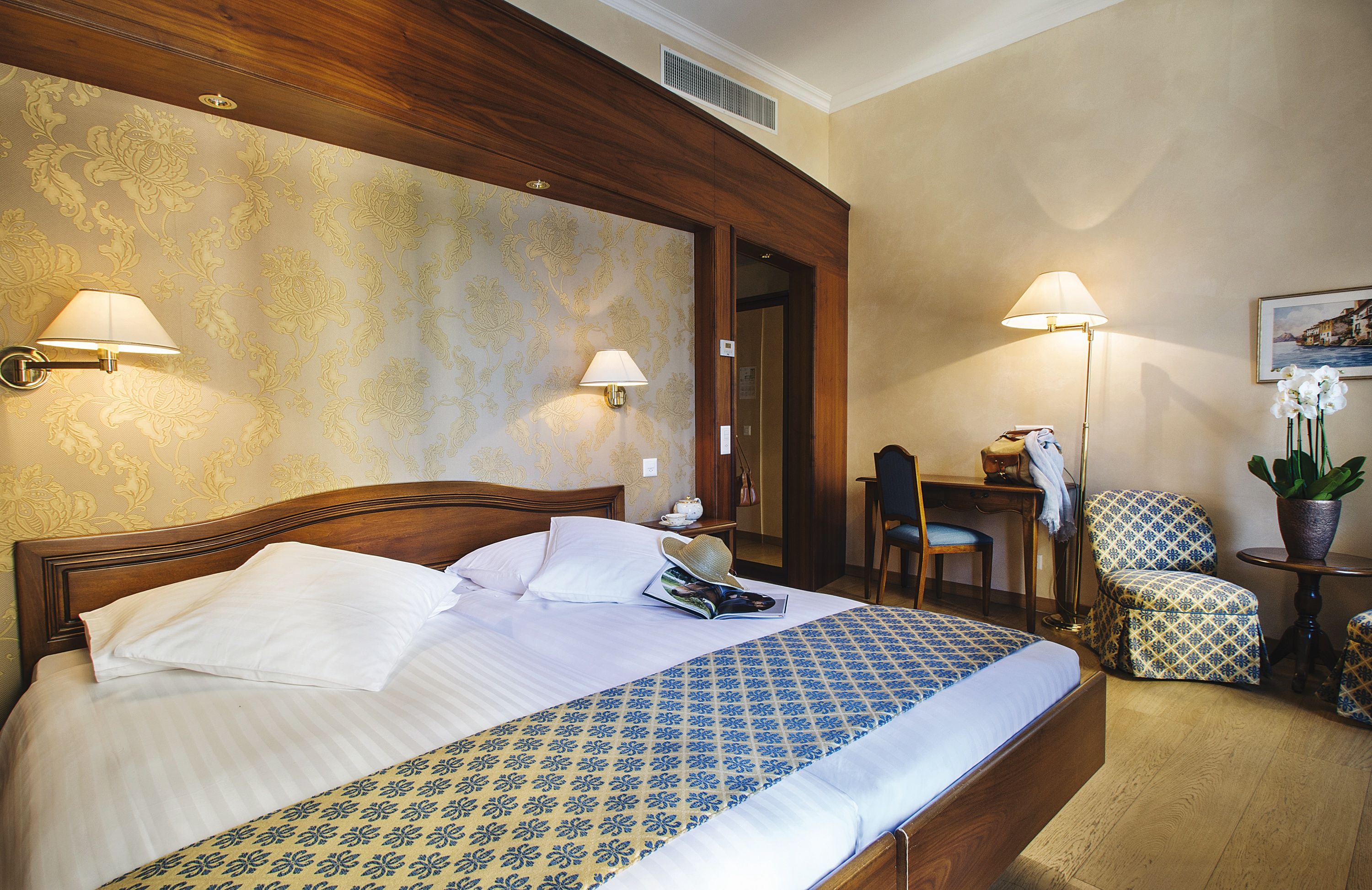 Quarto de hotel com uma cama grande, duas luminárias de cabeceira, uma escrivaninha com cadeira e um vaso de planta.