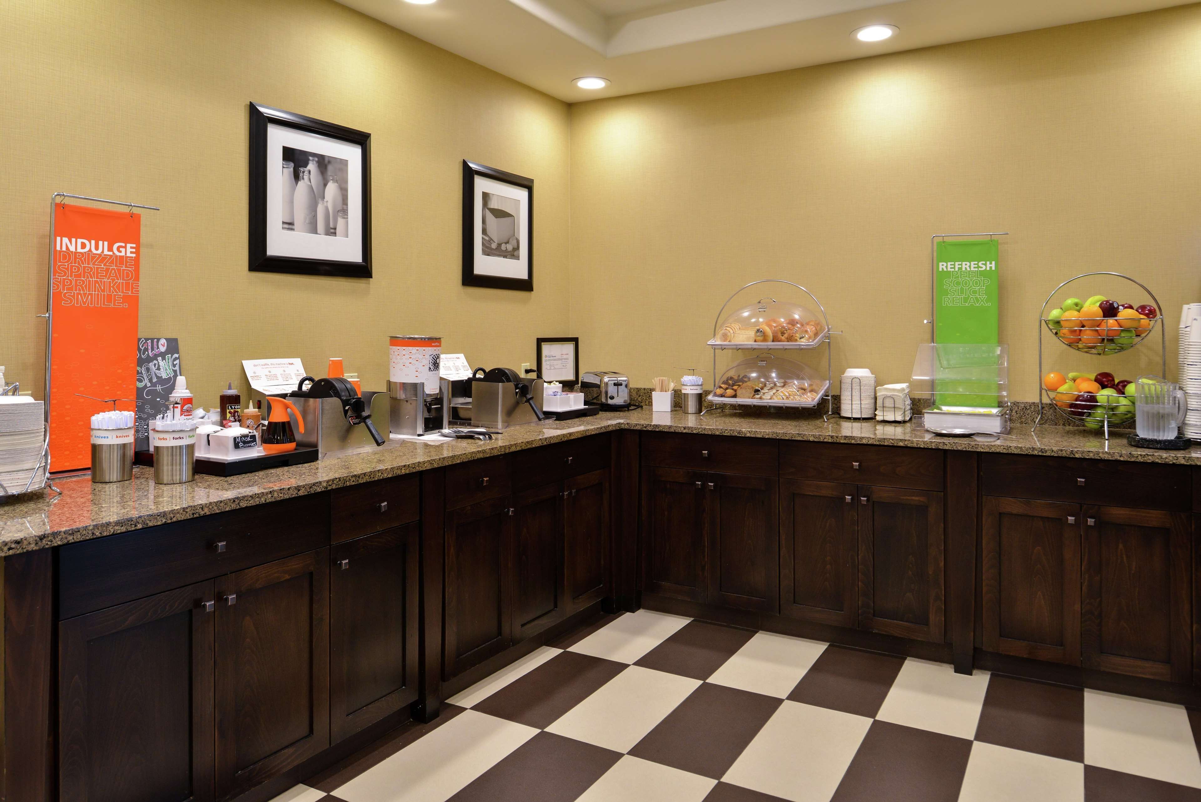 Cozinha de hotel com chão xadrez, armários de madeira, um espaço para café e uma apresentação de frutas.