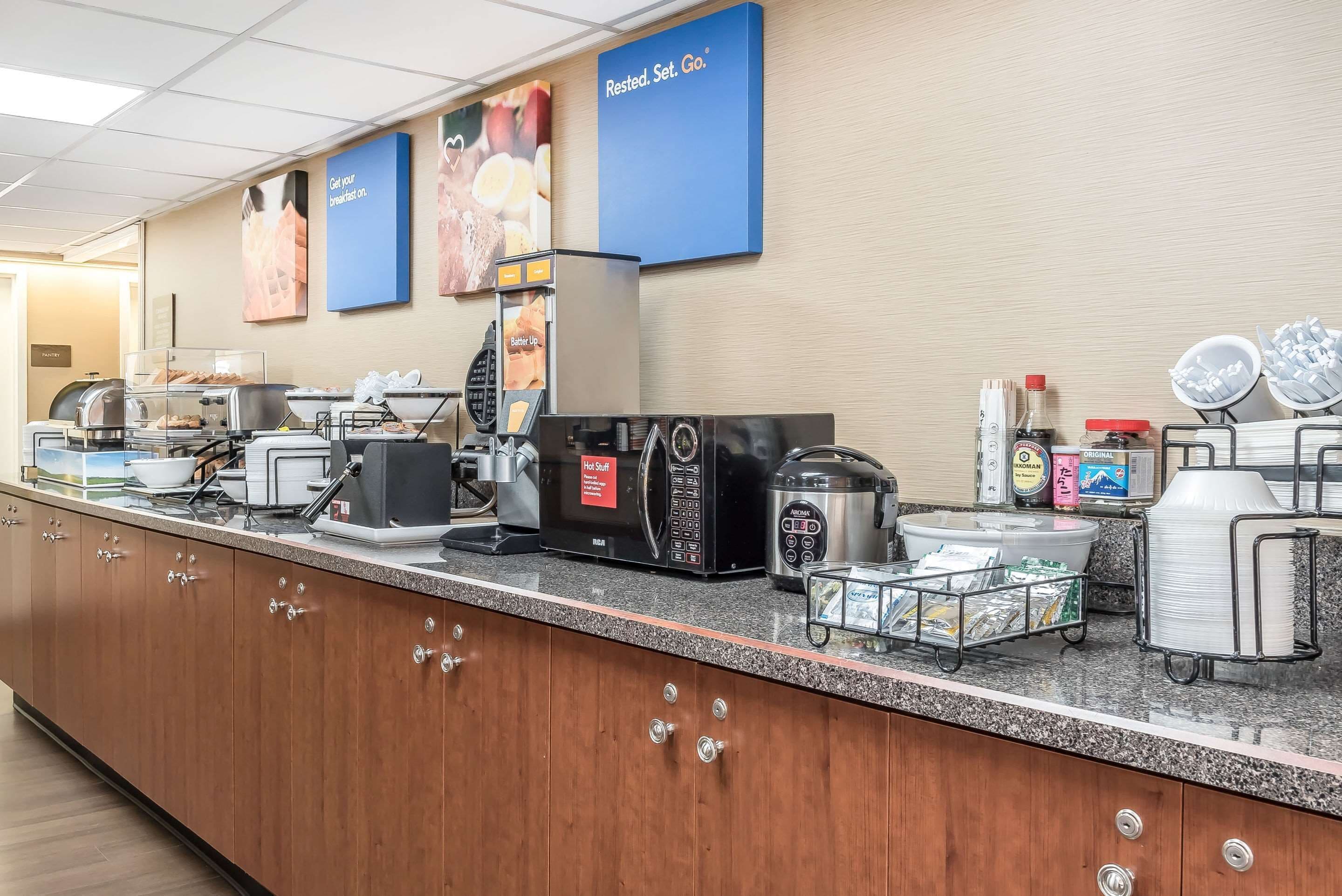 Espaço de café da manhã em hotel, com serviço de buffet, incluindo máquina de café, estação de comidas quentes e diversos recipientes com alimentos.