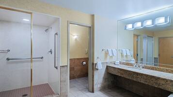 Casa de banho de hotel com cabine de duche em vidro, toucador com bancada de granito e espelho grande com iluminação superior.