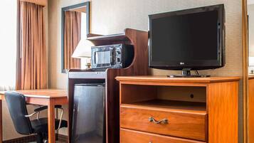 Quarto de hotel com uma escrivaninha de madeira, televisão, mini-geladeira, cadeira e cômoda de madeira com gavetas.