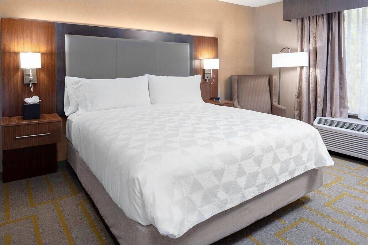 Quarto de hotel com uma cama grande, uma mesinha de cabeceira com abajur, uma cadeira, uma janela com cortinas e um aquecedor fixado na parede.