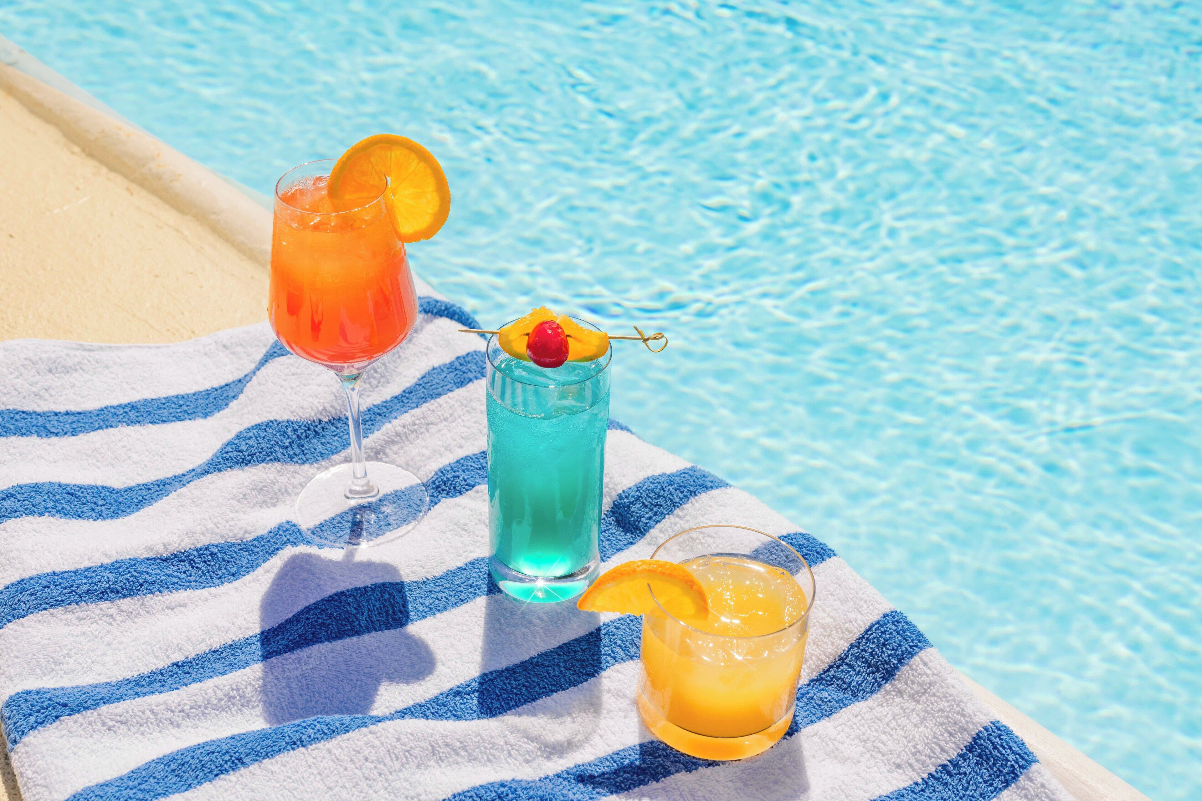 Três bebidas sobre uma toalha listrada à beira da piscina.