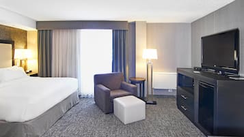 Quarto de hotel com uma cama grande, uma poltrona cinza, um banquinho pequeno, uma mesa de trabalho com uma TV de tela plana e duas luminárias.