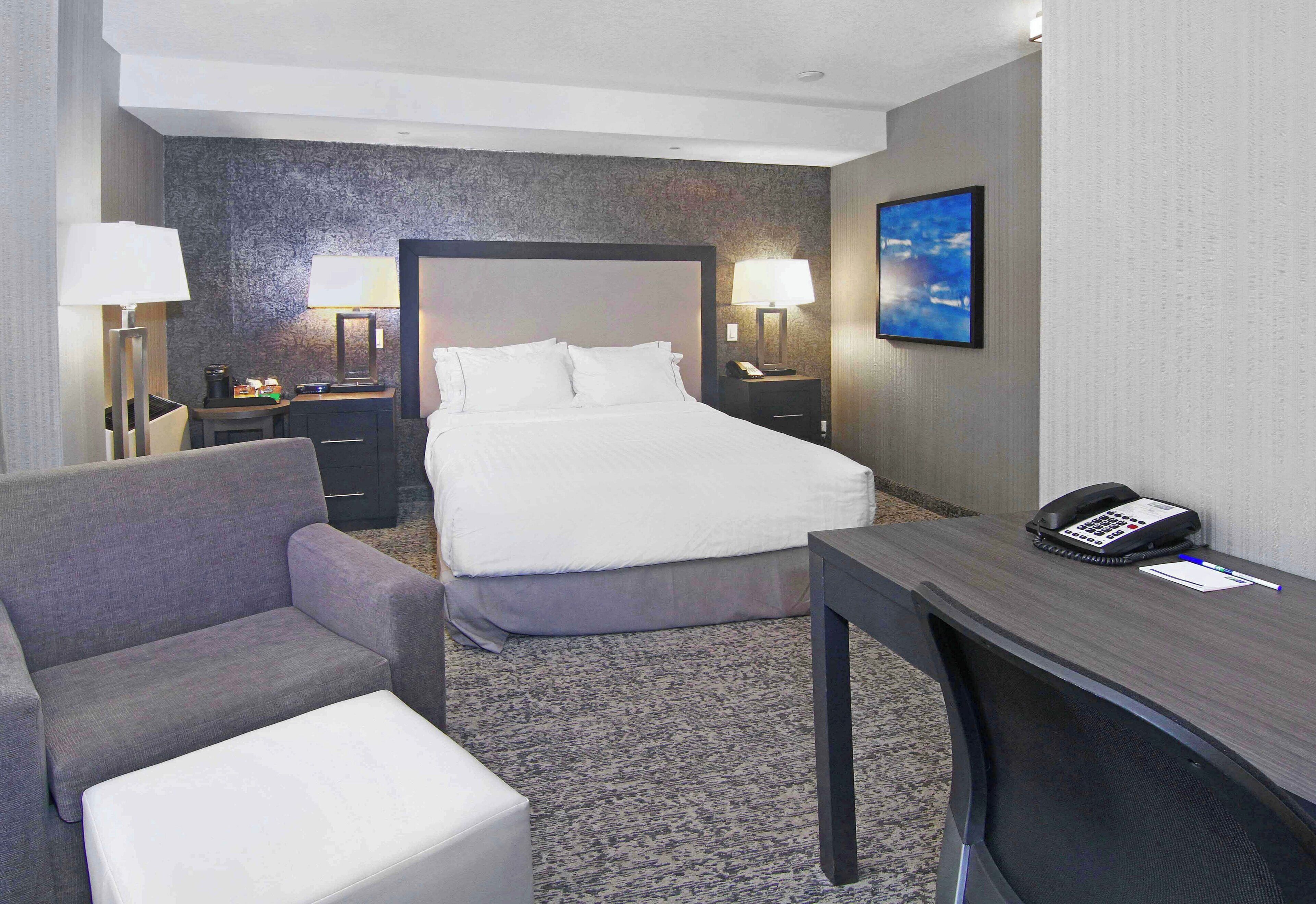 Quarto de hotel com uma cama, escrivaninha, cadeira, mesa de cabeceira e luminária.