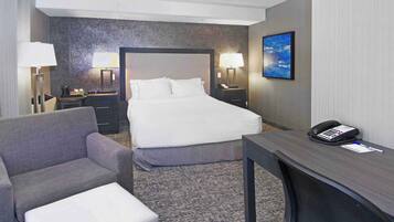 Quarto de hotel com uma cama, escrivaninha, cadeira, mesa de cabeceira e luminária.