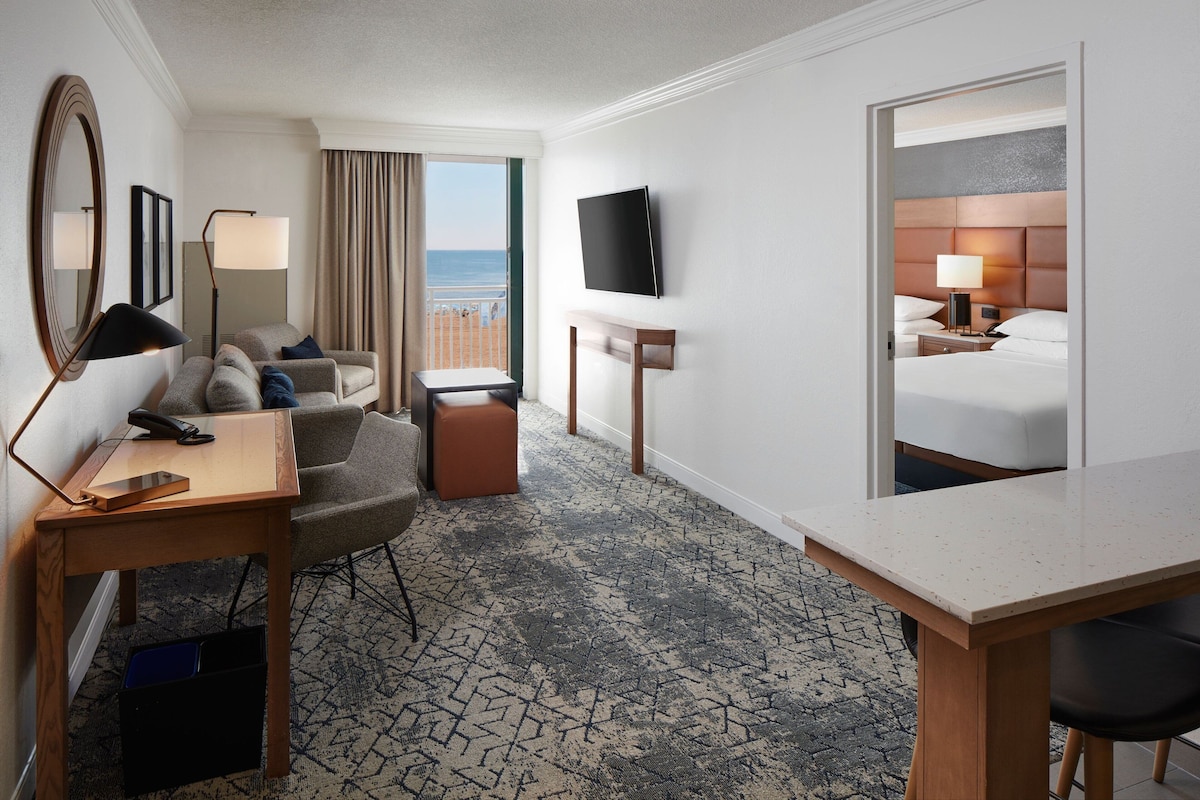 Quarto de hotel com cama, escrivaninha, cadeira, televisão, espelho e vista para a praia.