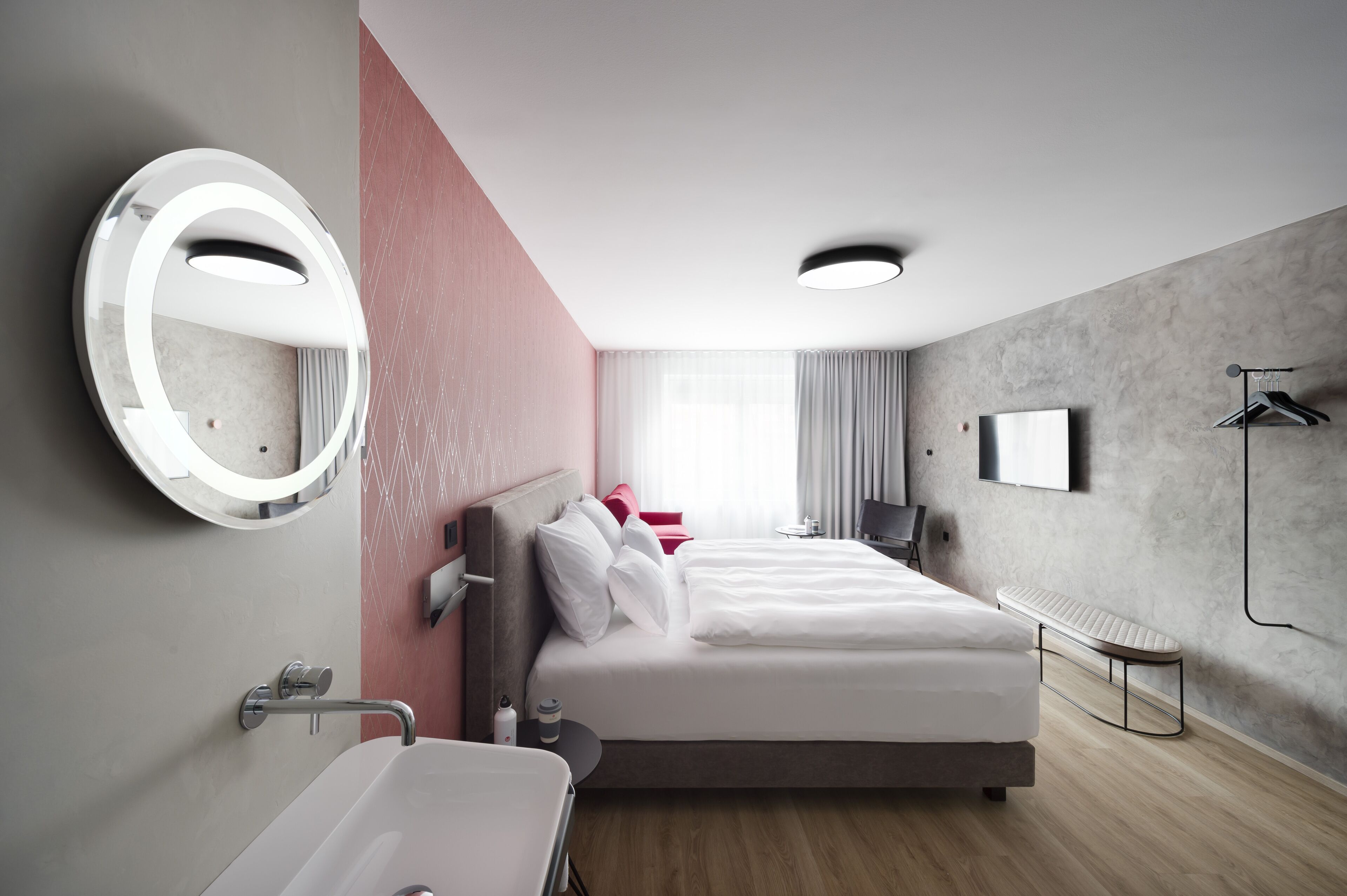 Quarto de hotel moderno com uma cama grande, um espelho redondo, uma mesinha de cabeceira, uma TV e um espaço com chuveiro.