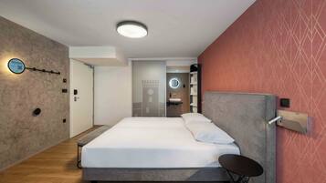 Quarto de hotel moderno com uma cama grande, uma mesa de cabeceira, prateleira embutida e um banheiro visível através de uma porta aberta.