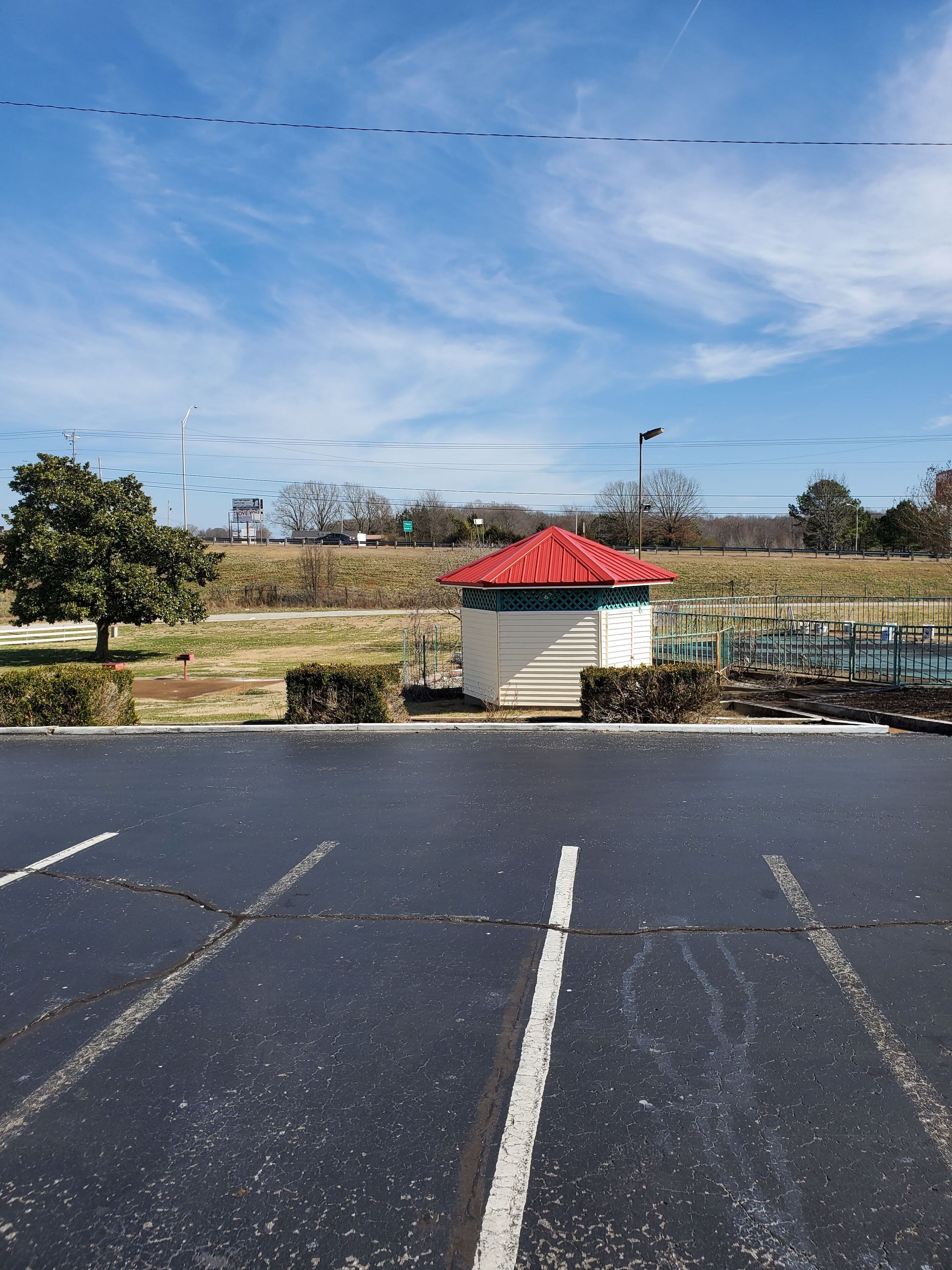Um parque de estacionamento com uma pequena construção de telhado vermelho, possivelmente um quiosque ou arrecadação, cercado por uma vedação.