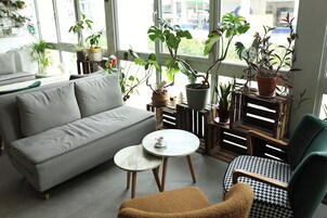 Uma sala de estar moderna com um sofá, uma mesa de centro e uma poltrona. Há várias plantas em vasos e uma vista do exterior por meio de janelas amplas.