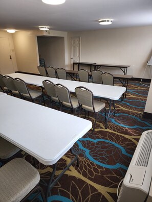 Sala de conferências com mesas e cadeiras, piso coberto de carpete e luminárias no teto.