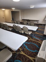 Sala de conferências com mesas e cadeiras, piso coberto de carpete e luminárias no teto.