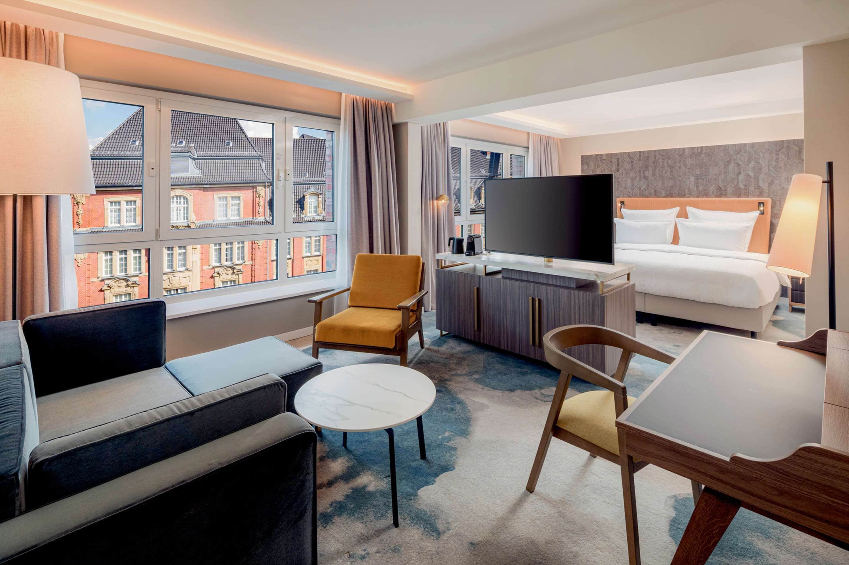 Quarto de hotel com sofá, poltrona, mesa de jantar, televisão e cama com cabeceira.