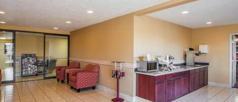 Um hall de hotel com mesa de recepção, micro-ondas, máquina de café e área de estar com cadeiras estampadas.