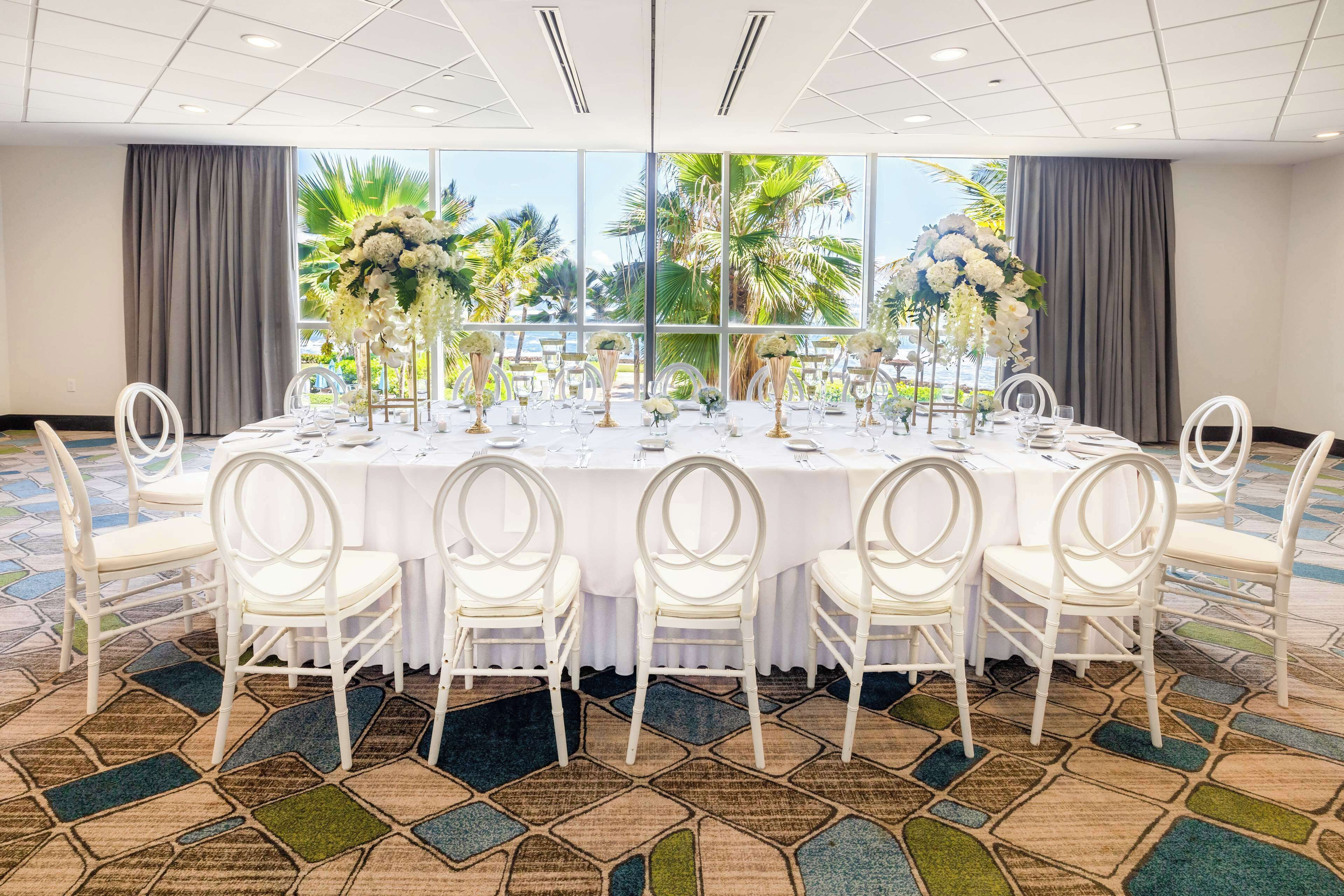 Uma sala de conferência com uma grande mesa redonda, cadeiras brancas e centros de mesa florais.