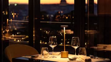 Mesa de jantar posta para dois, com taças de vinho e uma vela, com vista para o horizonte urbano ao pôr do sol.