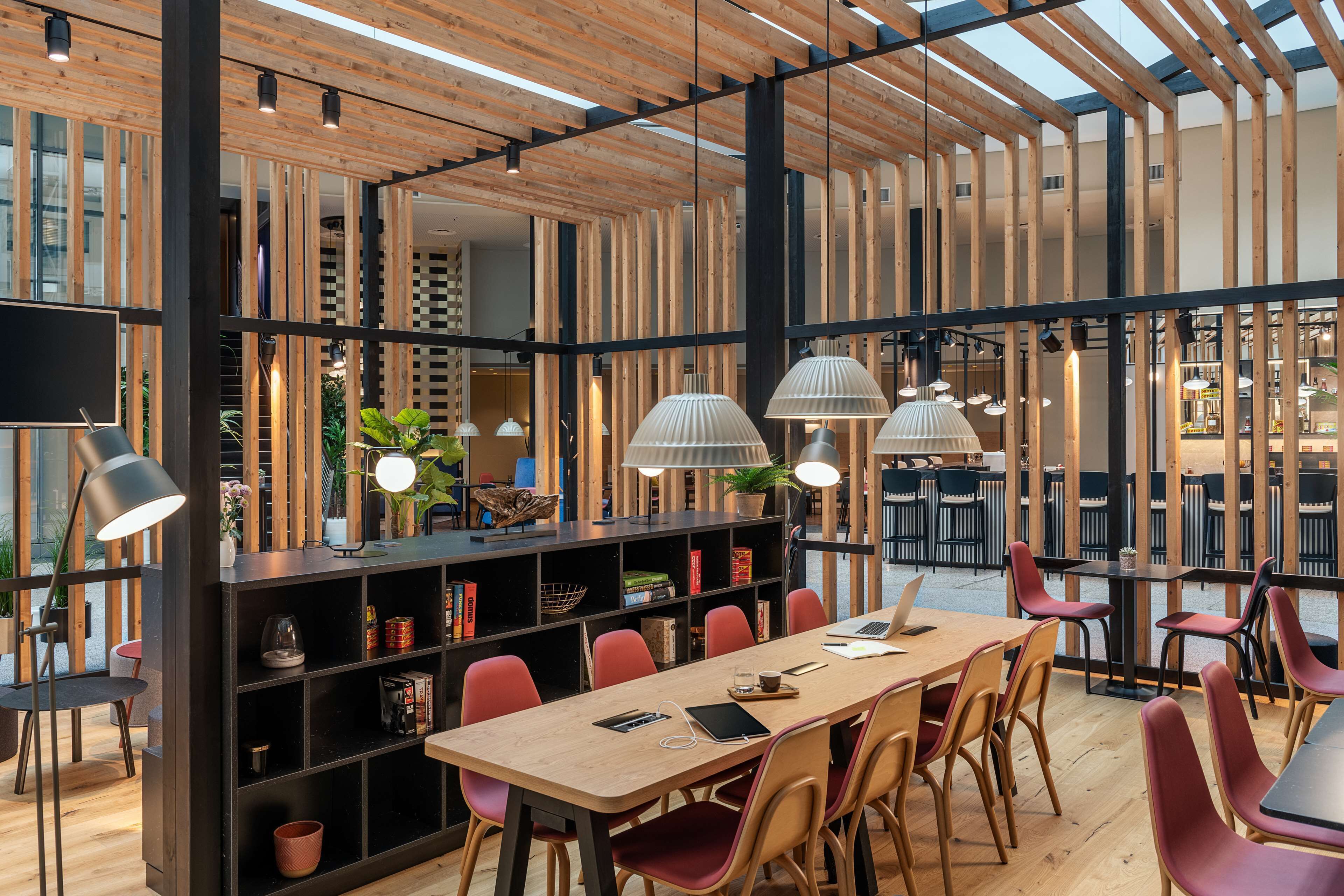 Um interior moderno com teto de madeira, estantes com moldura preta e cadeiras vermelhas ao redor de uma mesa central.