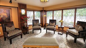 Quarto com quatro poltronas, um banco e uma mesa com abajur. Janelas grandes oferecem vista para o verde lá fora.