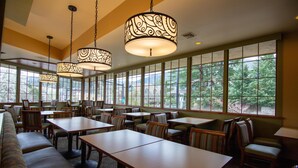 Um restaurante com várias mesas e cabines, amplas janelas com vista para árvores e luminárias decorativas pendentes.