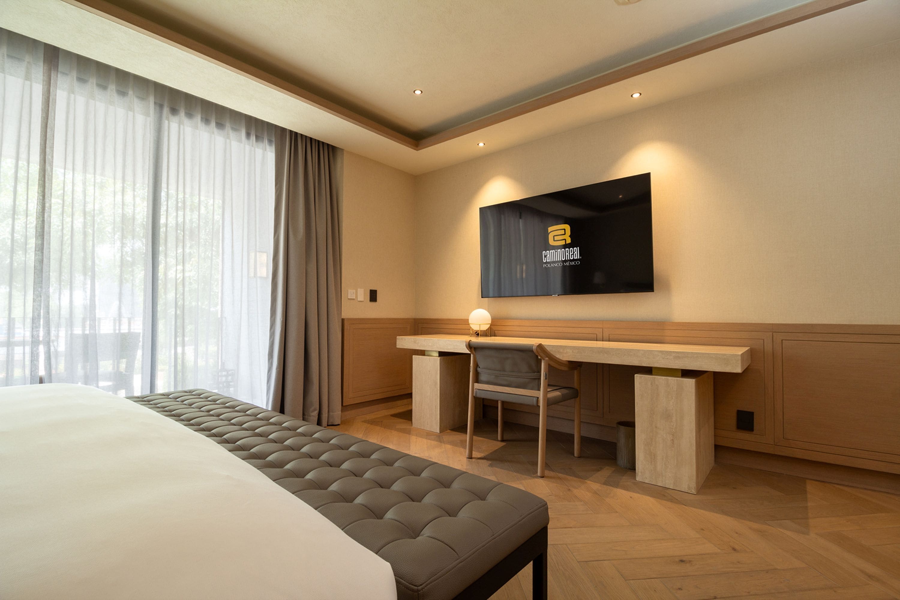 Quarto de hotel moderno com uma cama grande, uma escrivaninha com cadeira, televisão de tela plana na parede e uma janela com cortinas leves.