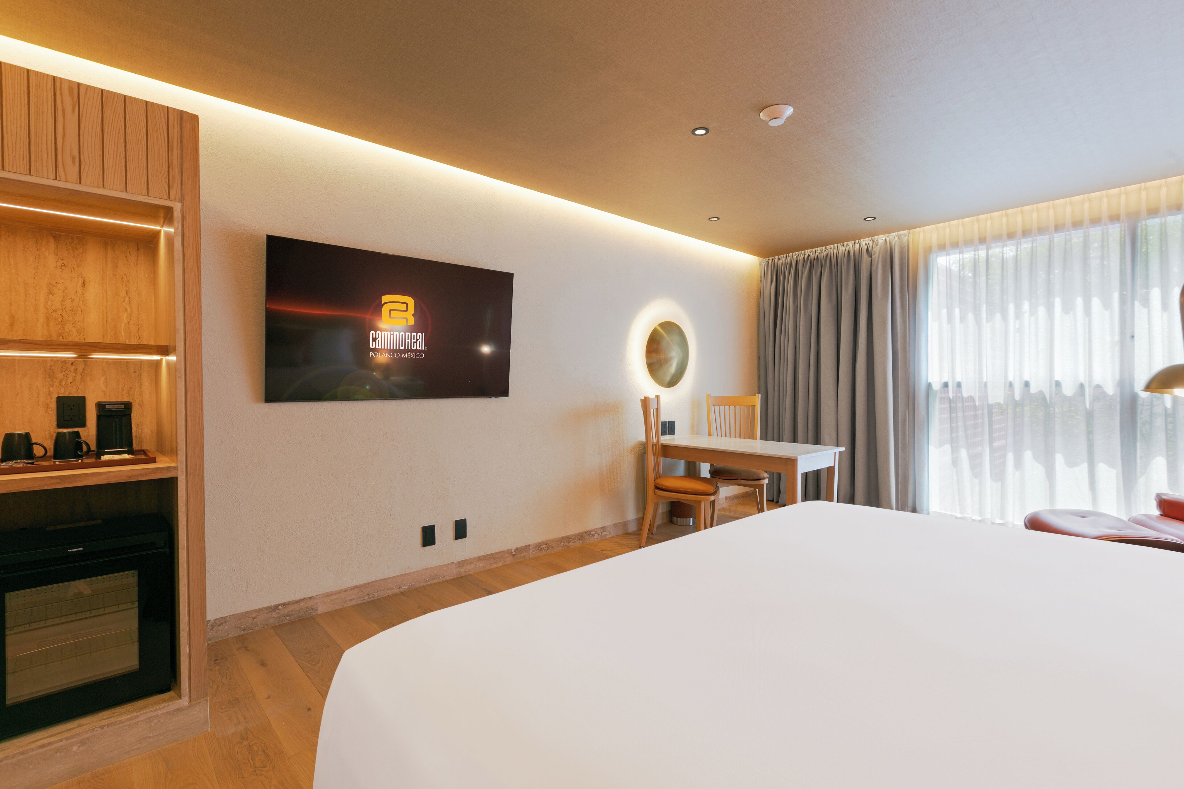 Quarto de hotel com cama, televisão na parede, escrivaninha com cadeira e janela com cortinas.