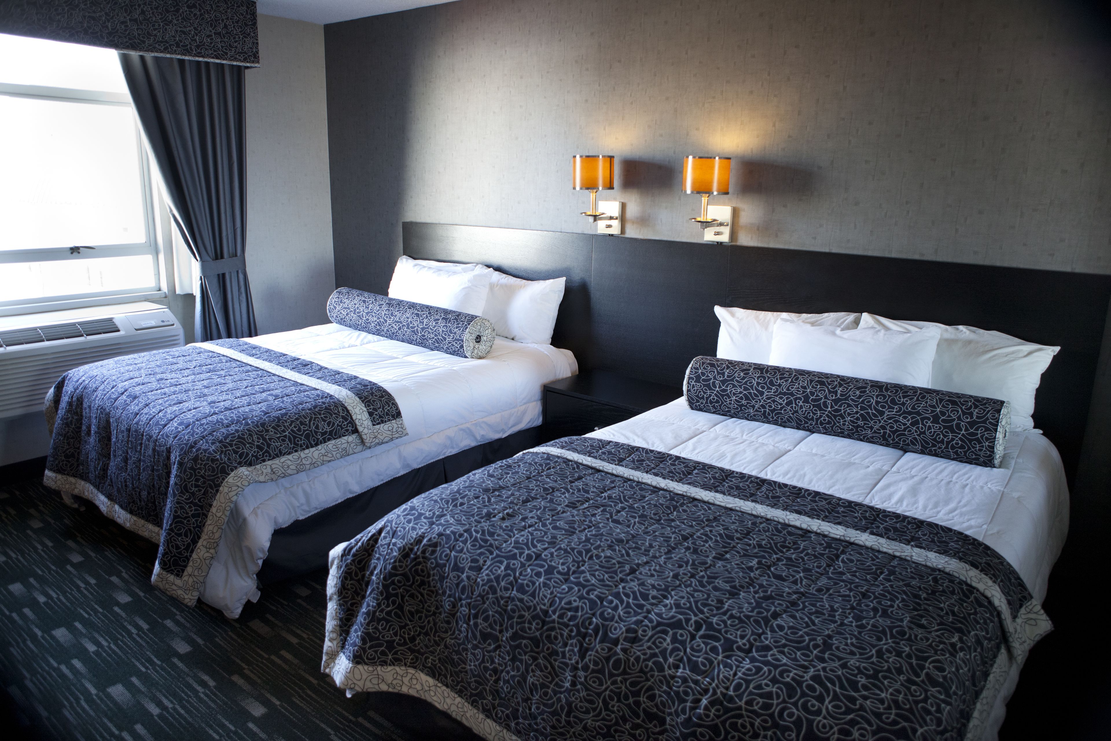 Quarto de hotel com duas camas, cabeceira escura e duas luminárias fixadas na parede.