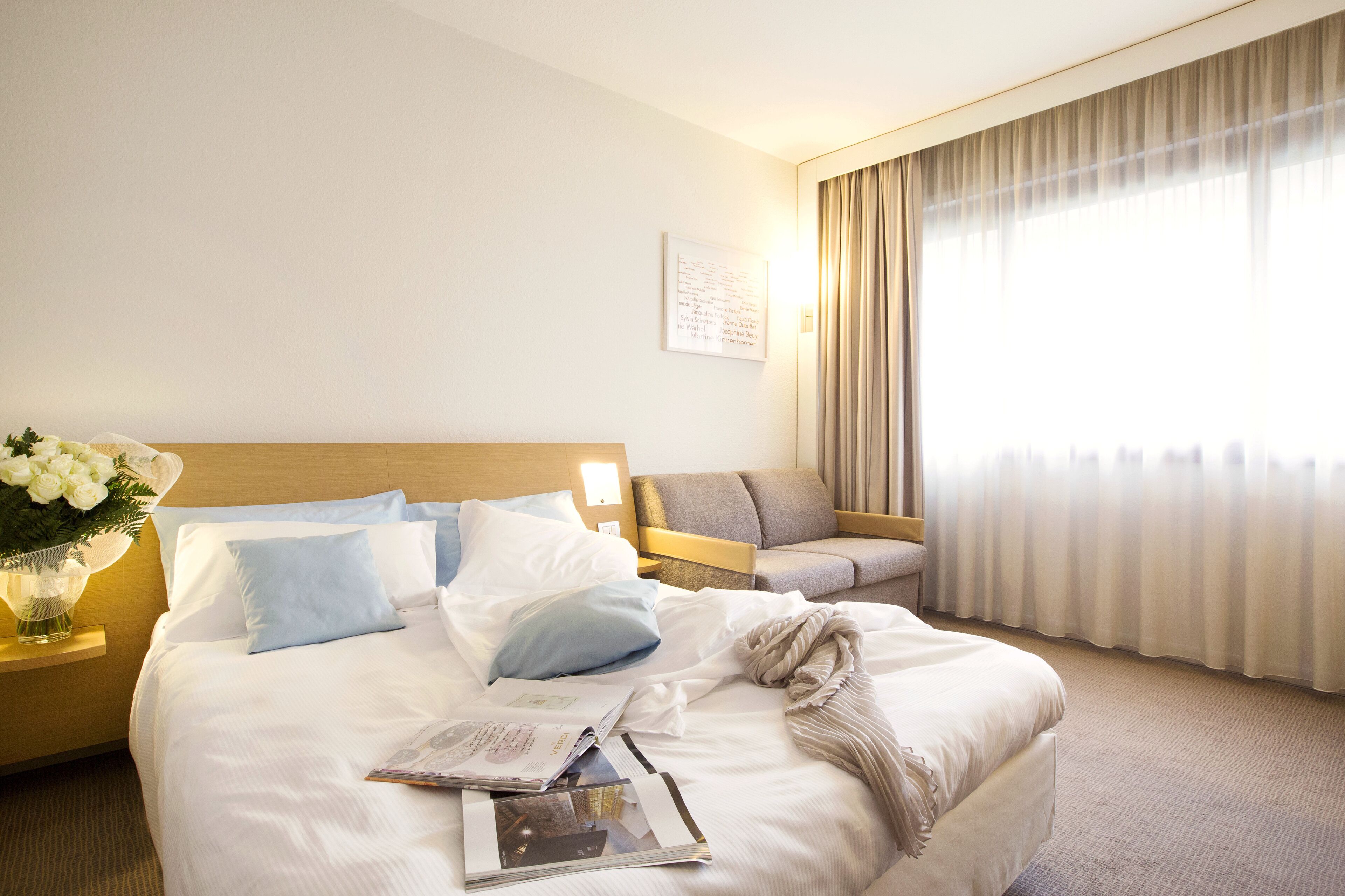Quarto de hotel com cama bem arrumada, sofá, mesa de cabeceira com luminária e janela com cortinas.