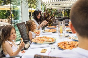Um grupo de pessoas jantando em uma mesa ao ar livre, com pizza, bebidas e um cardápio.