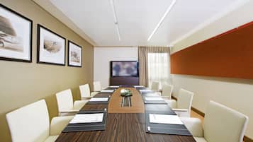 Uma sala de conferências com uma mesa comprida, cadeiras brancas, quadros nas paredes, uma televisão e uma parede de destaque vermelha.