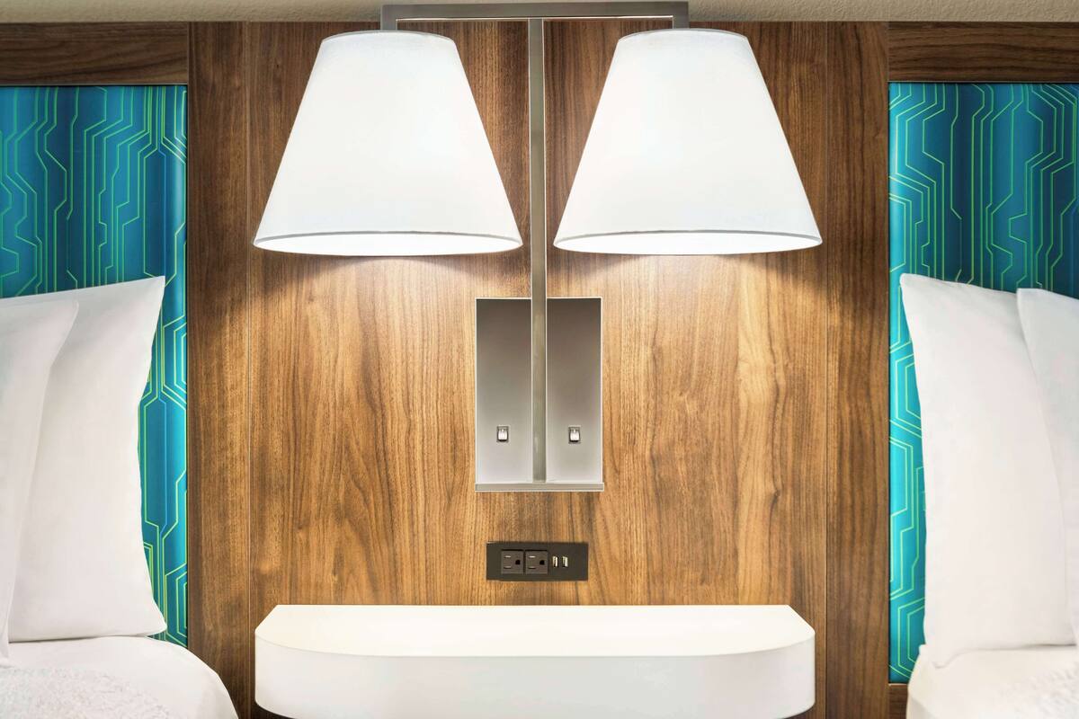 Quarto de hotel com cabeceira de madeira, duas luminárias de apoio, prateleira embutida e painel de controle com tomadas.