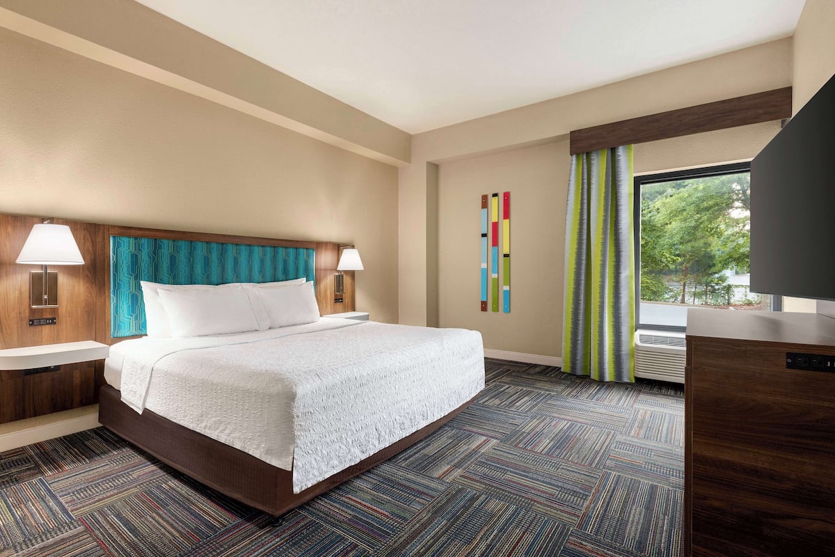 Quarto de hotel com uma cama grande, duas mesas de cabeceira com abajures, uma TV de tela plana, uma janela com cortinas e uma obra de arte abstrata colorida na parede.