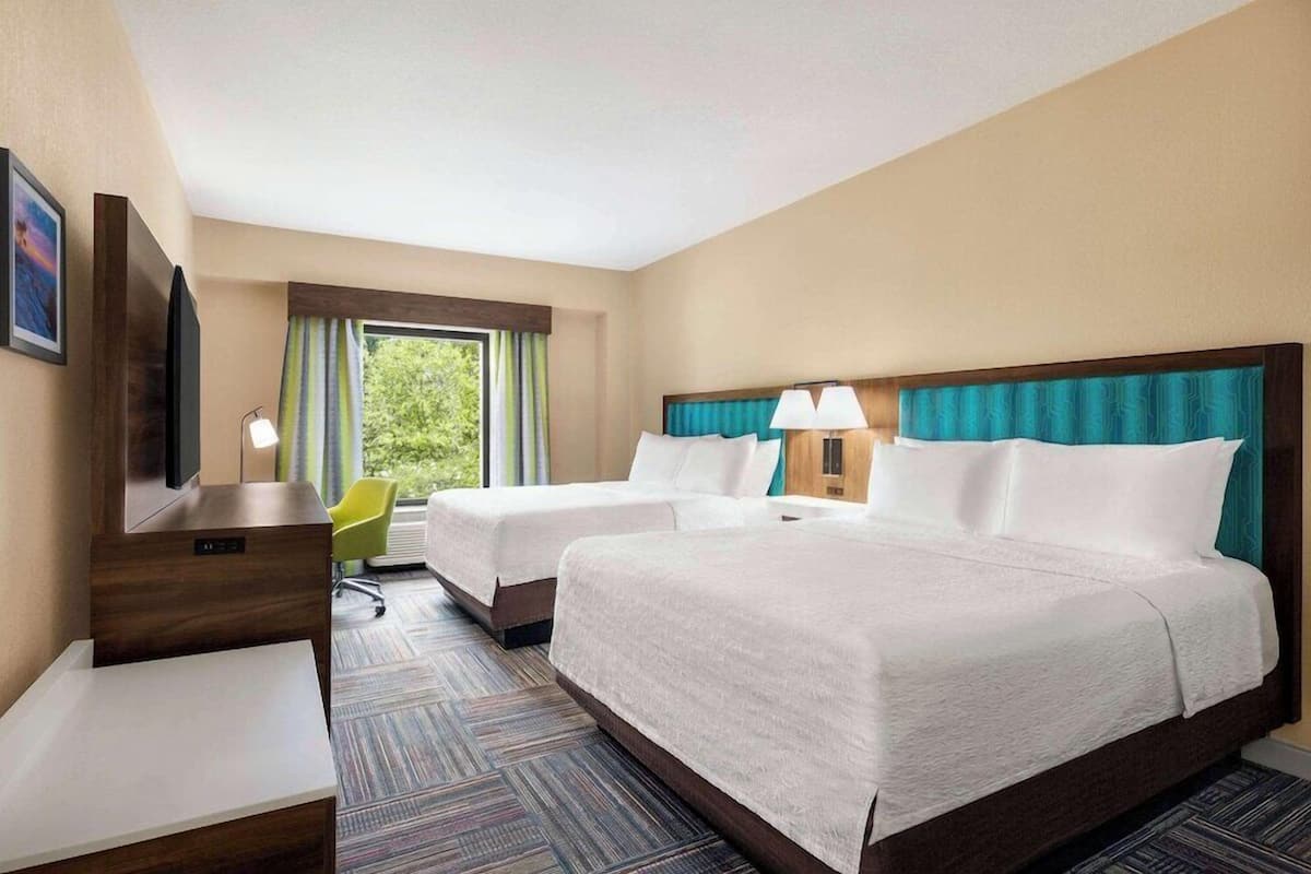 Quarto de hotel com duas camas, uma escrivaninha, uma cadeira, uma televisão e uma janela com cortinas.