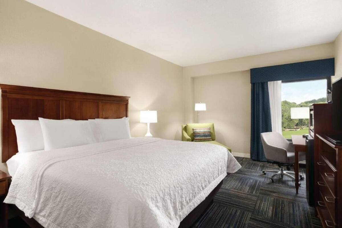 Quarto de hotel com uma cama grande, duas luminárias de cabeceira, uma mesa com cadeira, uma TV e vista para o exterior.