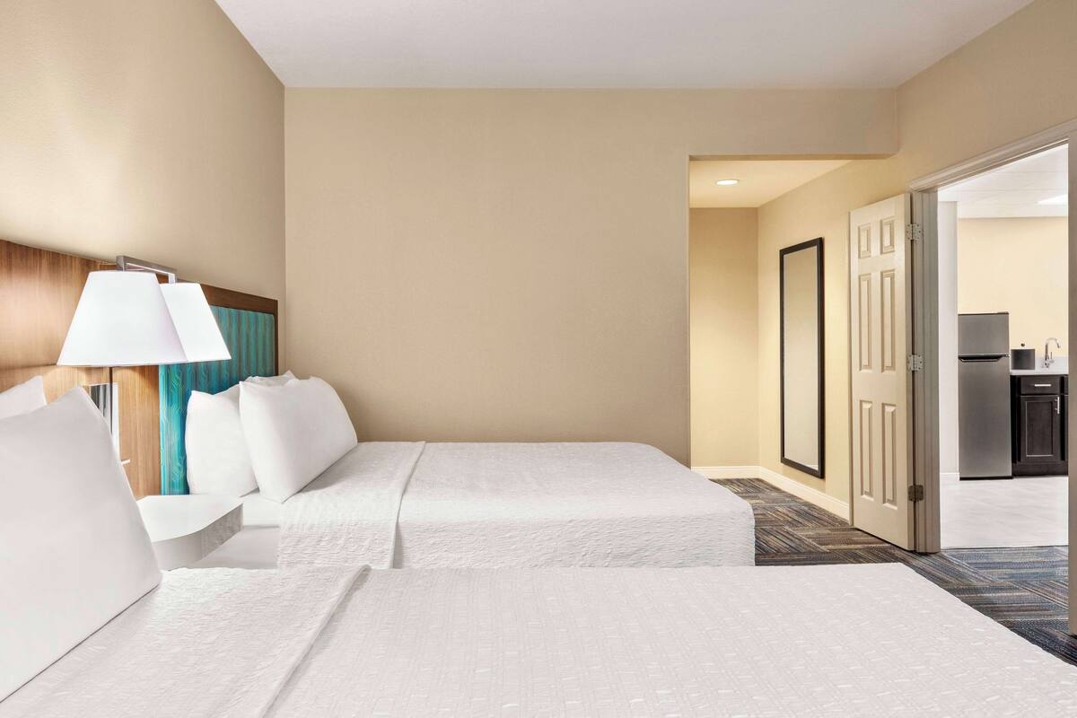 Quarto de hotel com duas camas, uma escrivaninha com luminária, porta para uma cozinha e vista para o corredor.