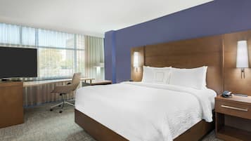 Quarto de hotel com uma cama grande, uma escrivaninha com televisão de tela plana, uma cadeira e uma janela com cortinas.